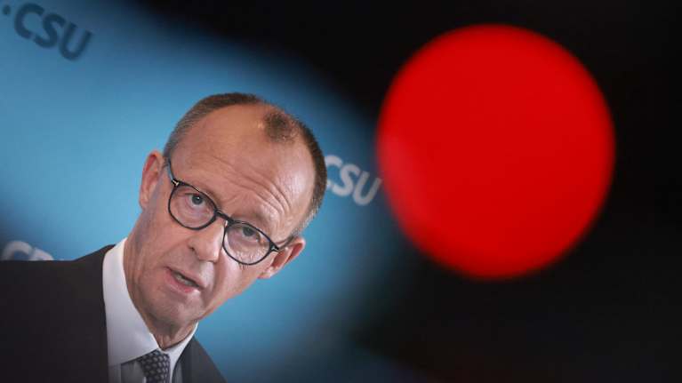 Vasemmalla vakava, silmälasipäinen mies katsoo kohti kameraa. Hänen takanaan näkyy seinällä CSU-kirjaimia ja hänen päänsä kohdalta oikealle seinällä heijastuu punainen pallo.