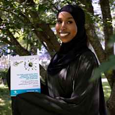 Idil Hussein seisoo puun siimeksessä kädessään väitöskirja, jota näyttää hymyillen kameralle. 