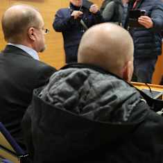 Kuvassa näkyvät henkilöt istuvat oikeussalissa. Taustalla valokuvaajat ottavat kuvia ja tekevät dokumentaatiota tilanteesta.