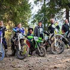 Seitsemän enduroharrastajaa kerääntyneenä yhteiskuvaan metsän reunaan. Kuvassa osalla ajovaatteet ja enduropyörät.
