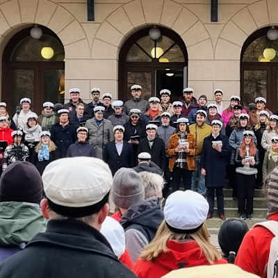 En kör sjunger på trappan till en stenbyggnad. Sångare och publik iklädda studentmössor.