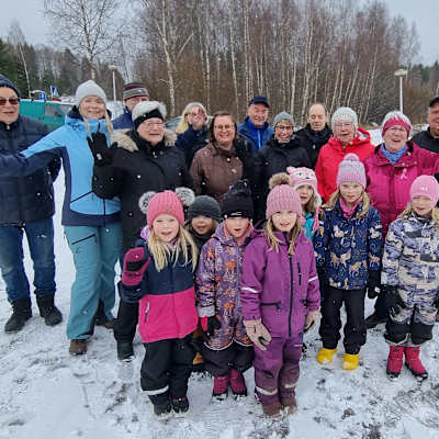 En grupp människor står tillsammans utomhus på en snötäckt mark, klädda i vinterkläder och tittar mot kameran. Det är en blandning av vuxna och barn, med träden och ett vintrigt landskap i bakgrunden.