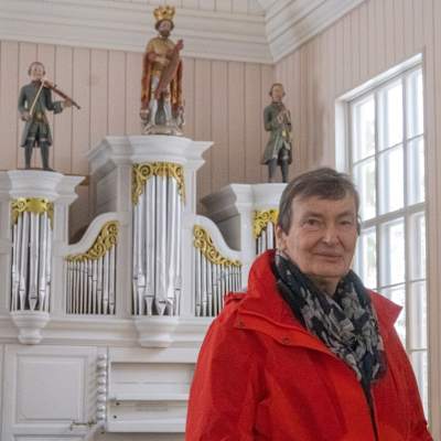 En kvinna i röd jacka står framför en gammal, sirligt utsmyckad kyrkorgel.